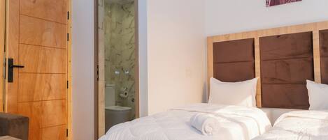 Quarto de hotel com guarda-roupa de madeira, casa de banho com cabine de duche em vidro e cama com roupa de cama branca.