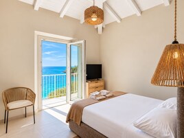 Quarto com cama, televisão, cadeira e vista para o mar através de uma porta de vidro.