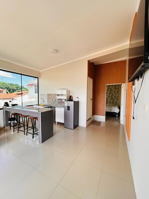 Cozinha moderna com ilha central, área de refeições com banquetas, geladeira e quarto com cama e lambrisagem de madeira.