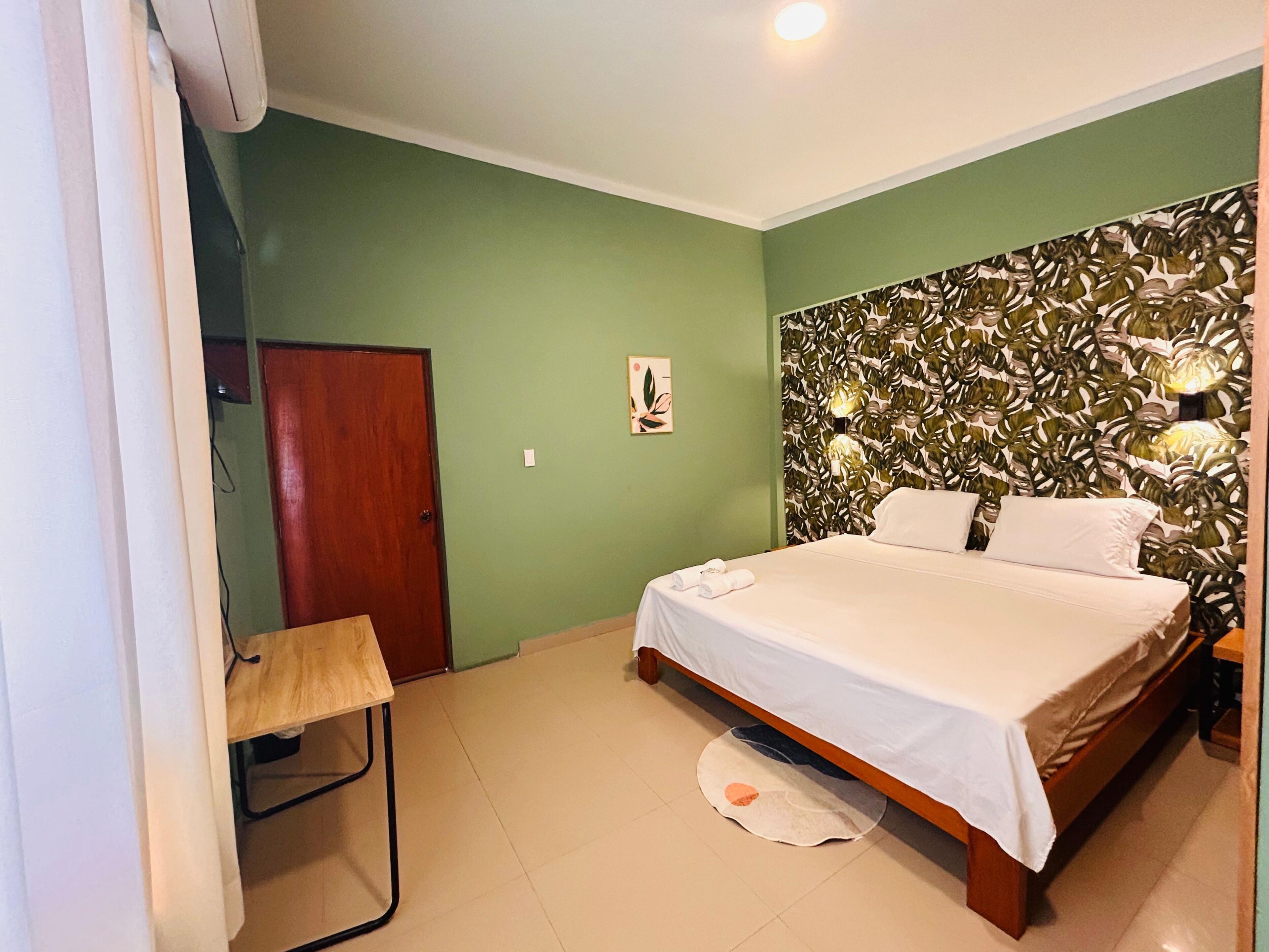 Quarto com cama, mesa de cabeceira, luminária na parede e porta.