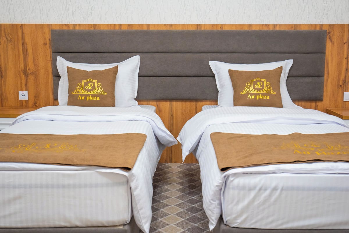 Quarto de hotel com duas camas, cada uma com cabeceira, almofada marrom com o logotipo "Air plaza" e manta marrom.