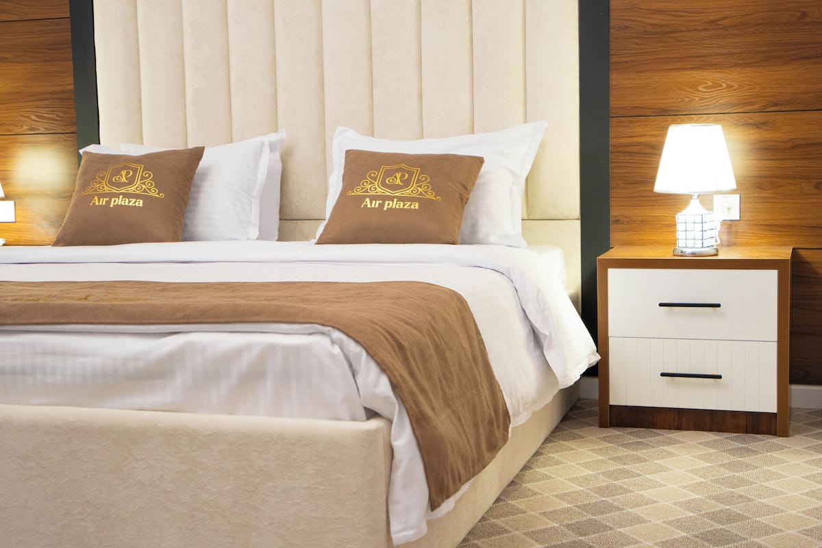 Uma cama bem arrumada com um cobertor marrom, duas almofadas com a marca 'Air plaza', um abajur e uma mesinha de cabeceira de madeira com gavetas.