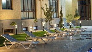 Cadeiras de descanso ao ar livre com painéis solares, piscina e um edifício com janelas amplas e iluminação.
