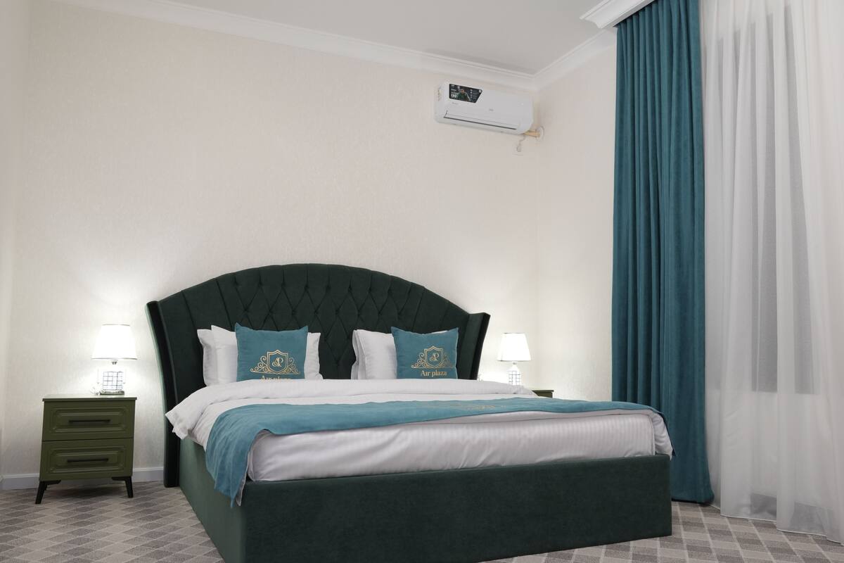 Cama bem arrumada com cabeceira escura, duas almofadas e manta azul-turquesa. Pequeno criado-mudo com abajur e objeto decorativo.