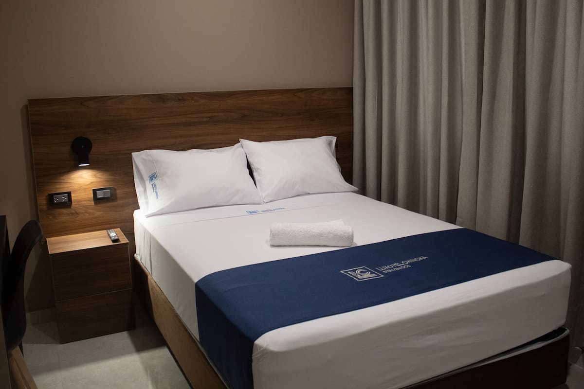 Quarto de hotel com cabeceira de madeira, roupa de cama branca, detalhe azul no cobreleito, mesa de cabeceira com controle remoto e mesa de apoio com abajur.