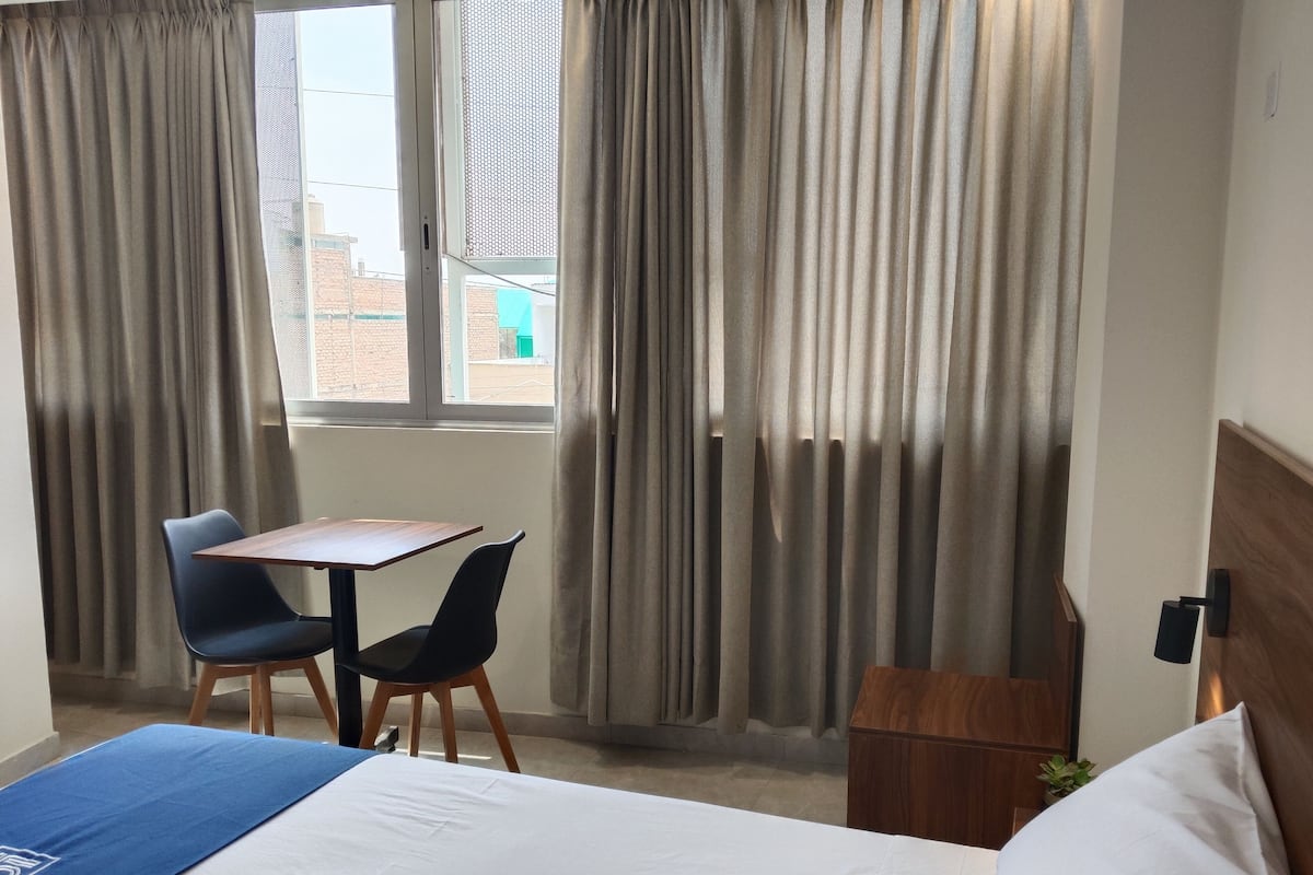Quarto de hotel com cama, uma mesa pequena, duas cadeiras e uma janela com cortinas.