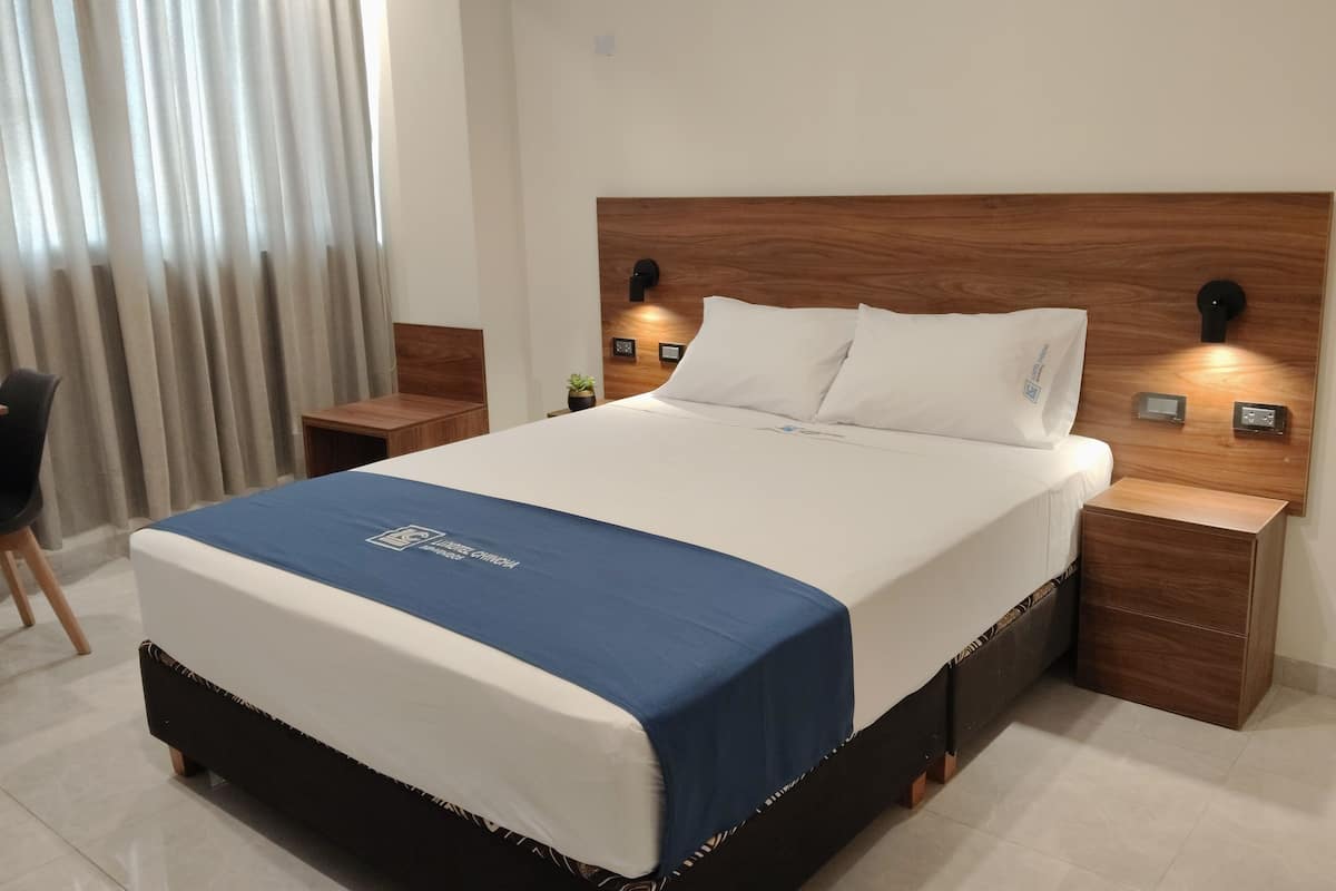 Quarto de hotel com uma cama grande, cabeceira de madeira, duas mesas de cabeceira e uma cadeira.