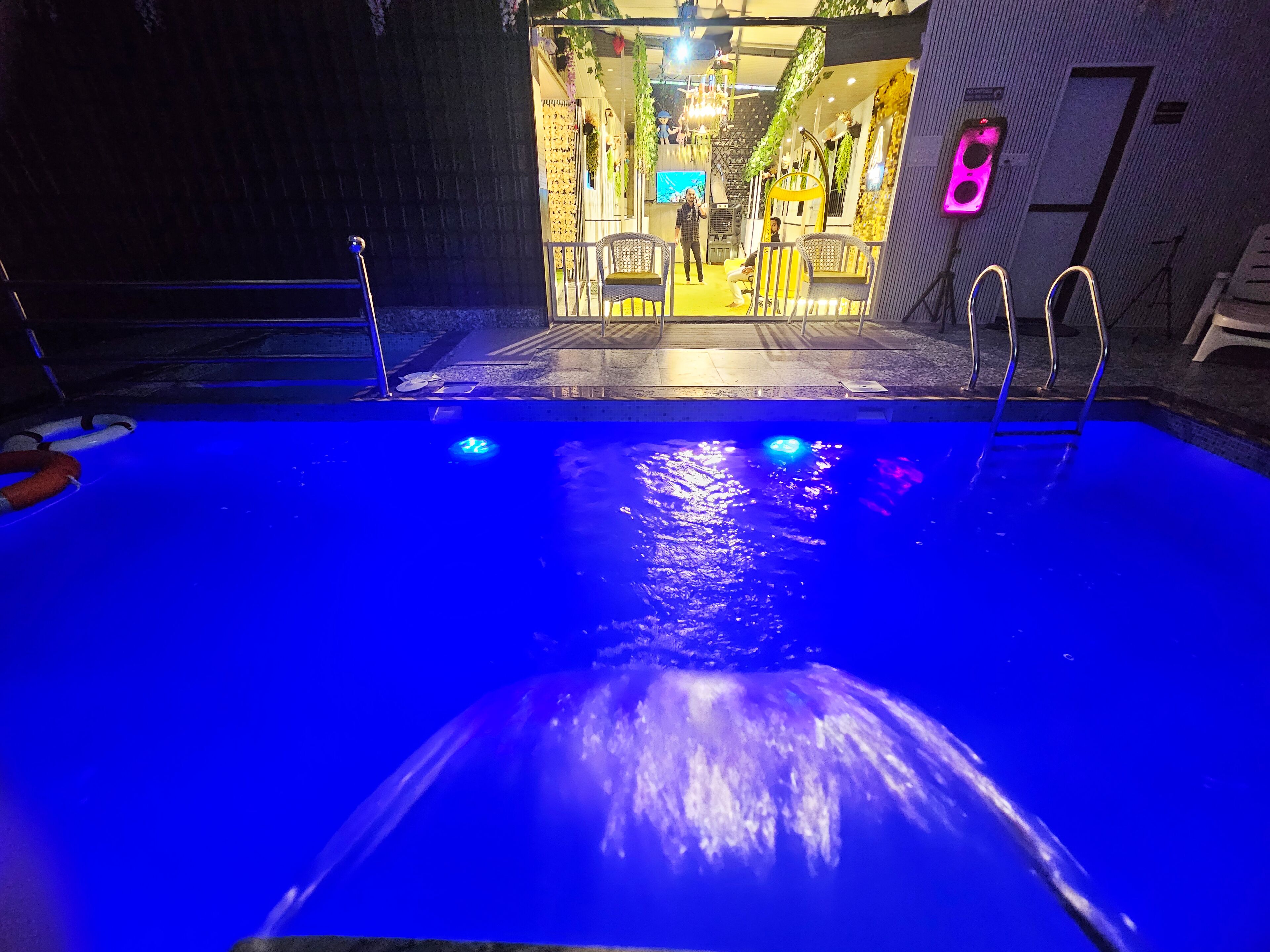 Piscina interna com superfície de água iluminada em azul, escada de metal e vista para área de entrada bem iluminada com cadeiras e uma pessoa ao fundo.