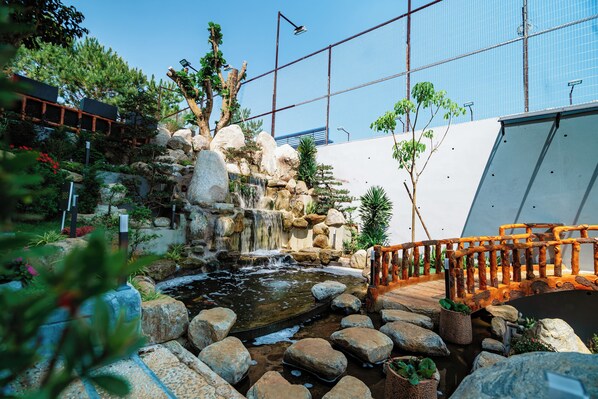 Um jardim de pedras com uma pequena cascata, cercado por diversas plantas e rochas.