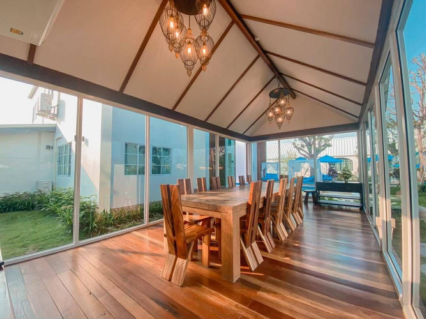 Um alpendre espaçoso com uma longa mesa de madeira e cadeiras, janelas amplas e um lustre pendurado.