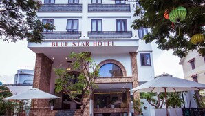 Exterior do Hotel Blue Star, com fachada de pedra, grandes janelas e área externa com mesas e guarda-sóis.