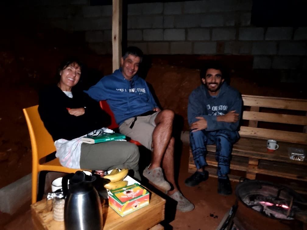 Três pessoas sentadas num banco em área externa com pouca luz, com uma parede de tijolos ao fundo.