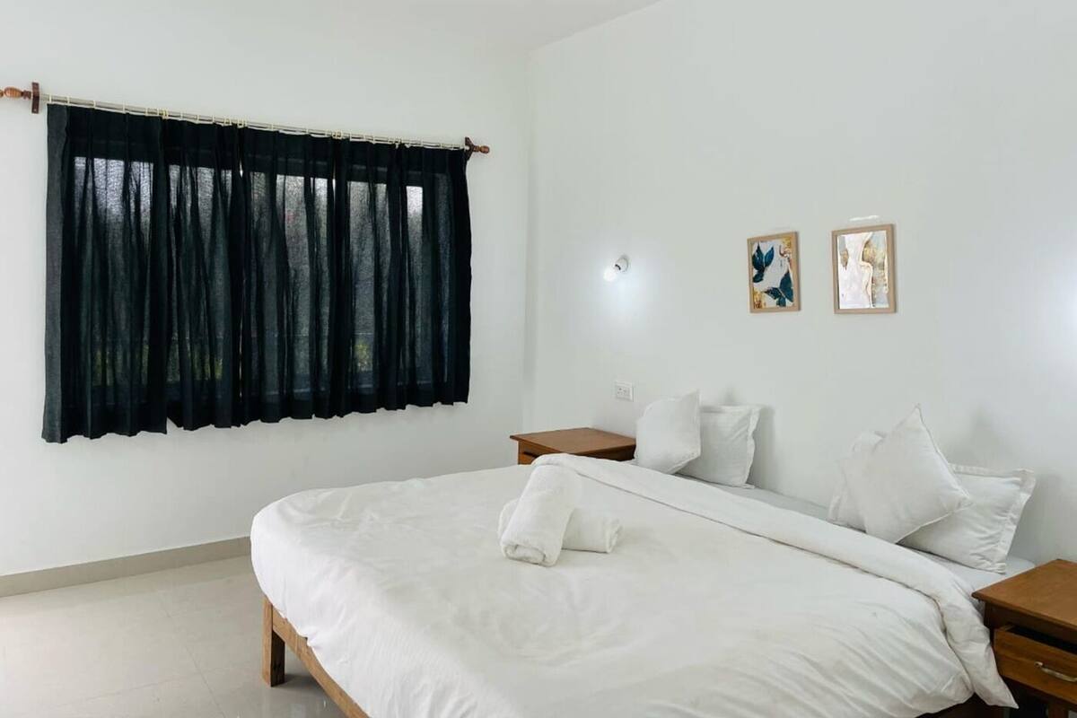 Quarto com uma cama grande, roupa de cama branca, uma mesinha de cabeceira de madeira, uma pequena mesa e duas luminárias fixadas na parede.