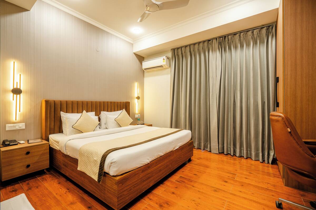Quarto de hotel com cama de madeira, luminárias de cabeceira, unidade de ar condicionado e uma grande janela com cortinas.
