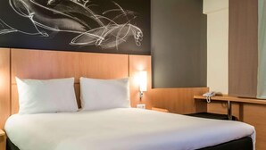 Quarto de hotel com uma cama grande, duas almofadas brancas, cabeceira de madeira, luminária fixada na parede e telefone sobre uma mesinha de madeira.