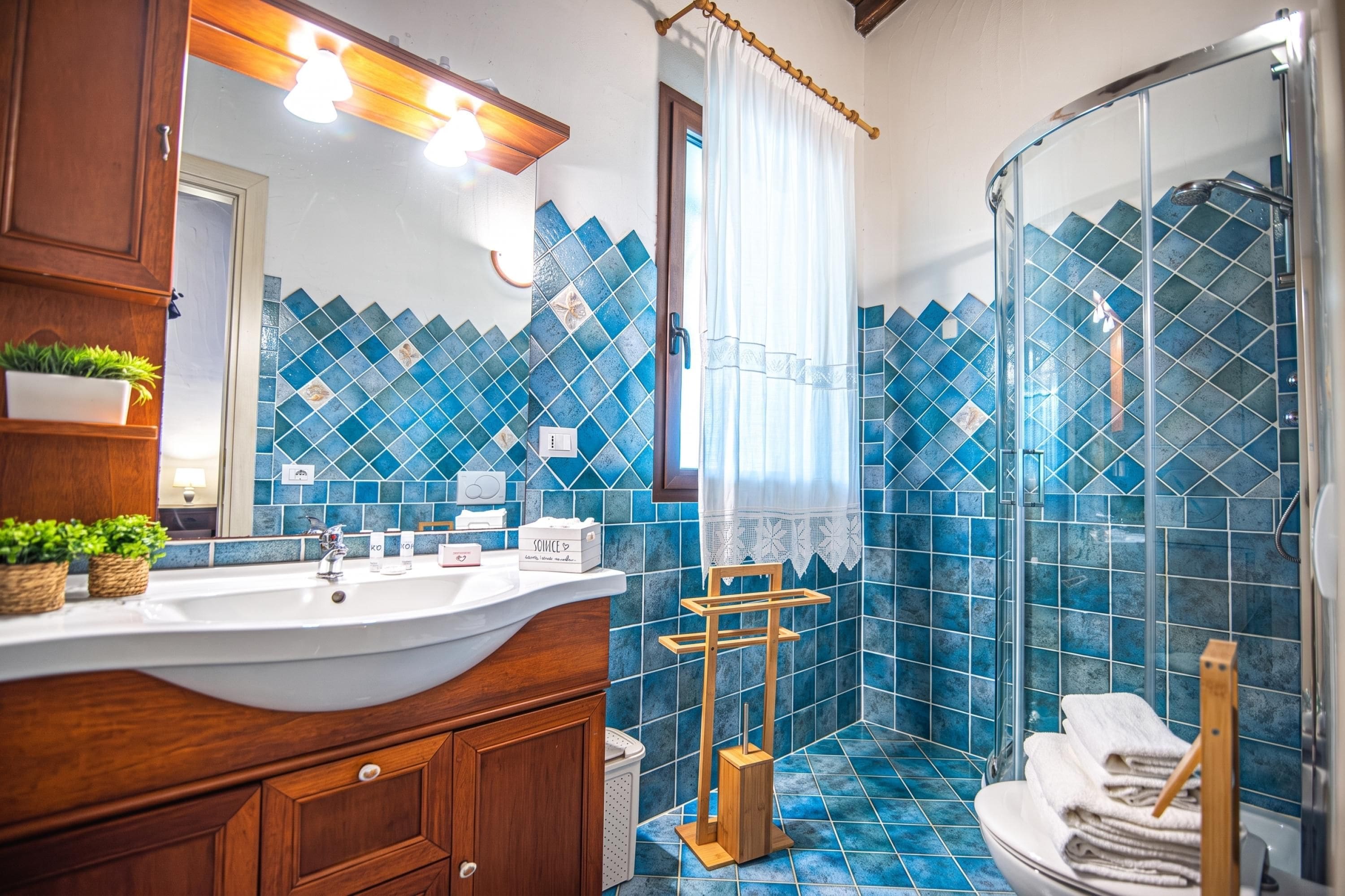 Casa de banho com toucador de madeira, lavatório e espelho. Existe uma área de duche com porta de vidro e azulejos azuis.