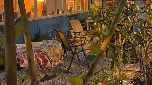 Uma casa com uma estrutura em forma de torre visível ao fundo, área de pátio com cadeiras e um jardim com plantas em vasos.