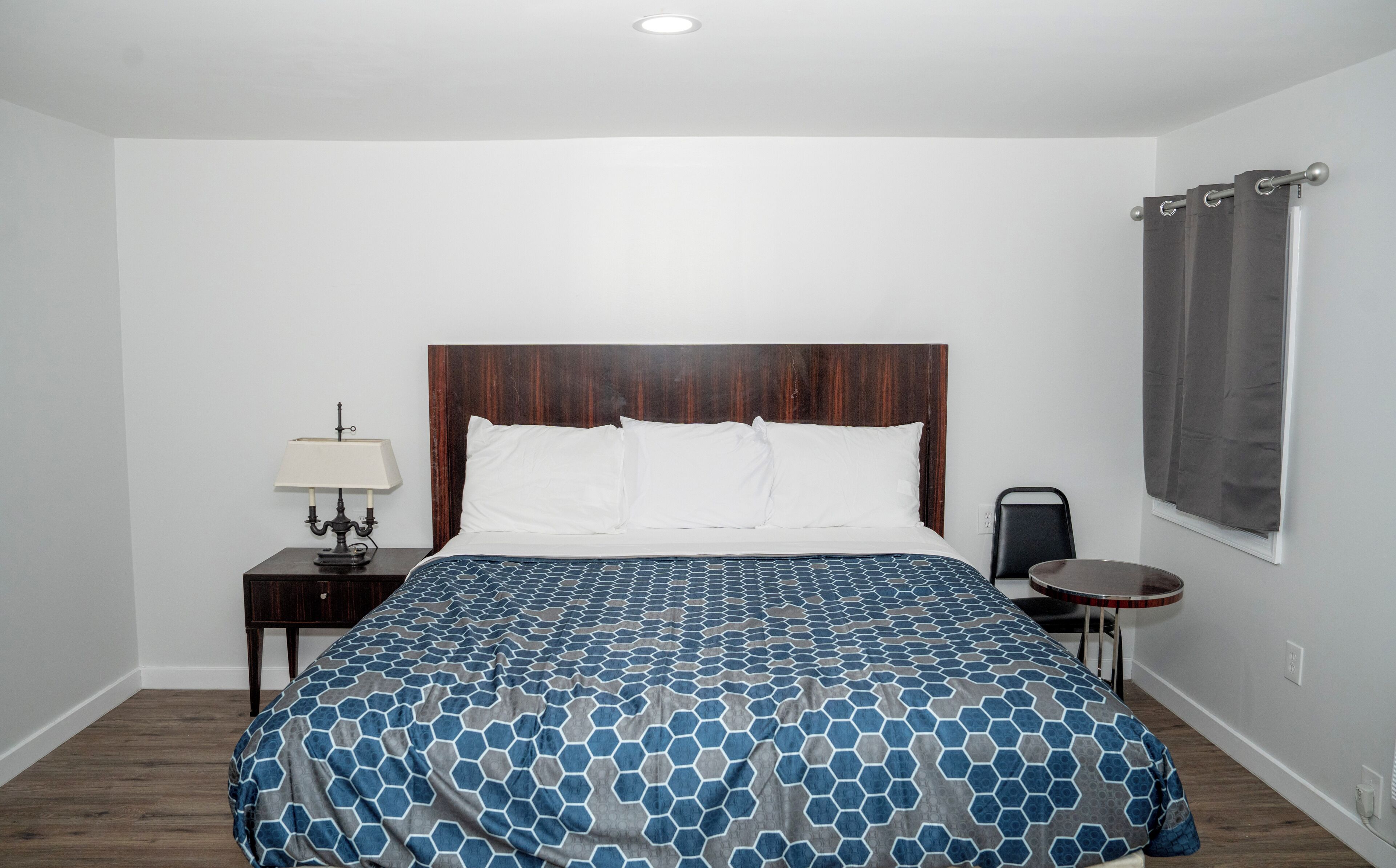 Quarto de hotel com cabeceira de madeira, cama com colcha azul e cinza estampada, mesa de cabeceira pequena com abajur, uma cadeira e uma janela com cortinas.