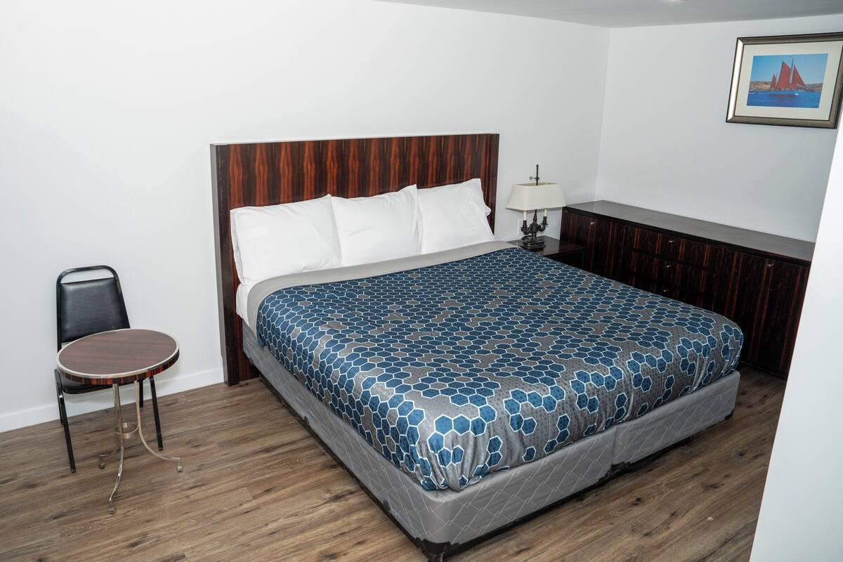 Quarto de hotel com uma cama grande, uma mesa redonda pequena, uma cadeira, uma mesinha de cabeceira com abajur e um quadro na parede.