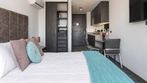 Quarto de hotel moderno com uma cama, uma escrivaninha e uma cadeira. Há um guarda-roupa e uma televisão fixada na parede.
