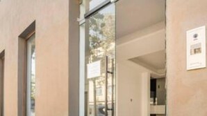 Entrada de um edifício moderno com porta de vidro, piso ladrilhado e uma placa visível na parede.