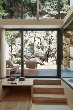 Um interior moderno com vista para um terreno rochoso através de grandes janelas, um banco de madeira e uma pequena mesa com um livro e uma tigela.