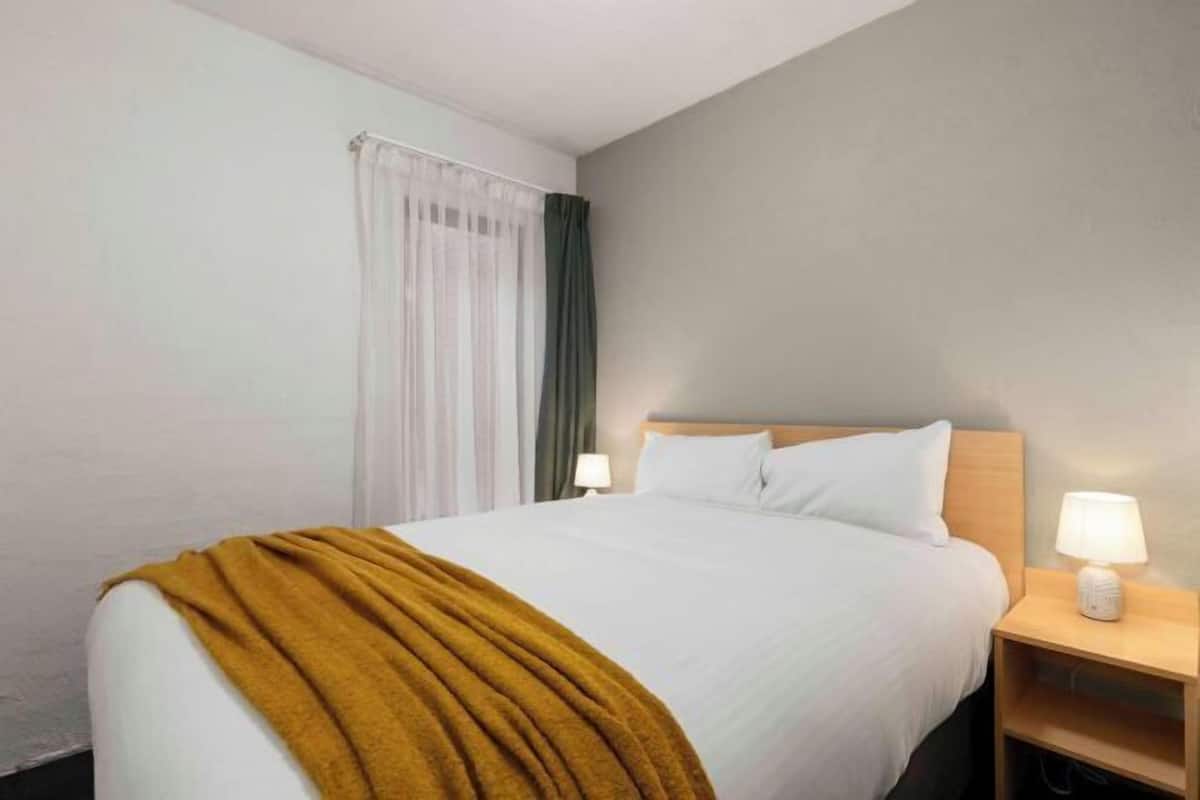 Quarto de hotel com cama, mesa de cabeceira com abajur e janela com cortinas.