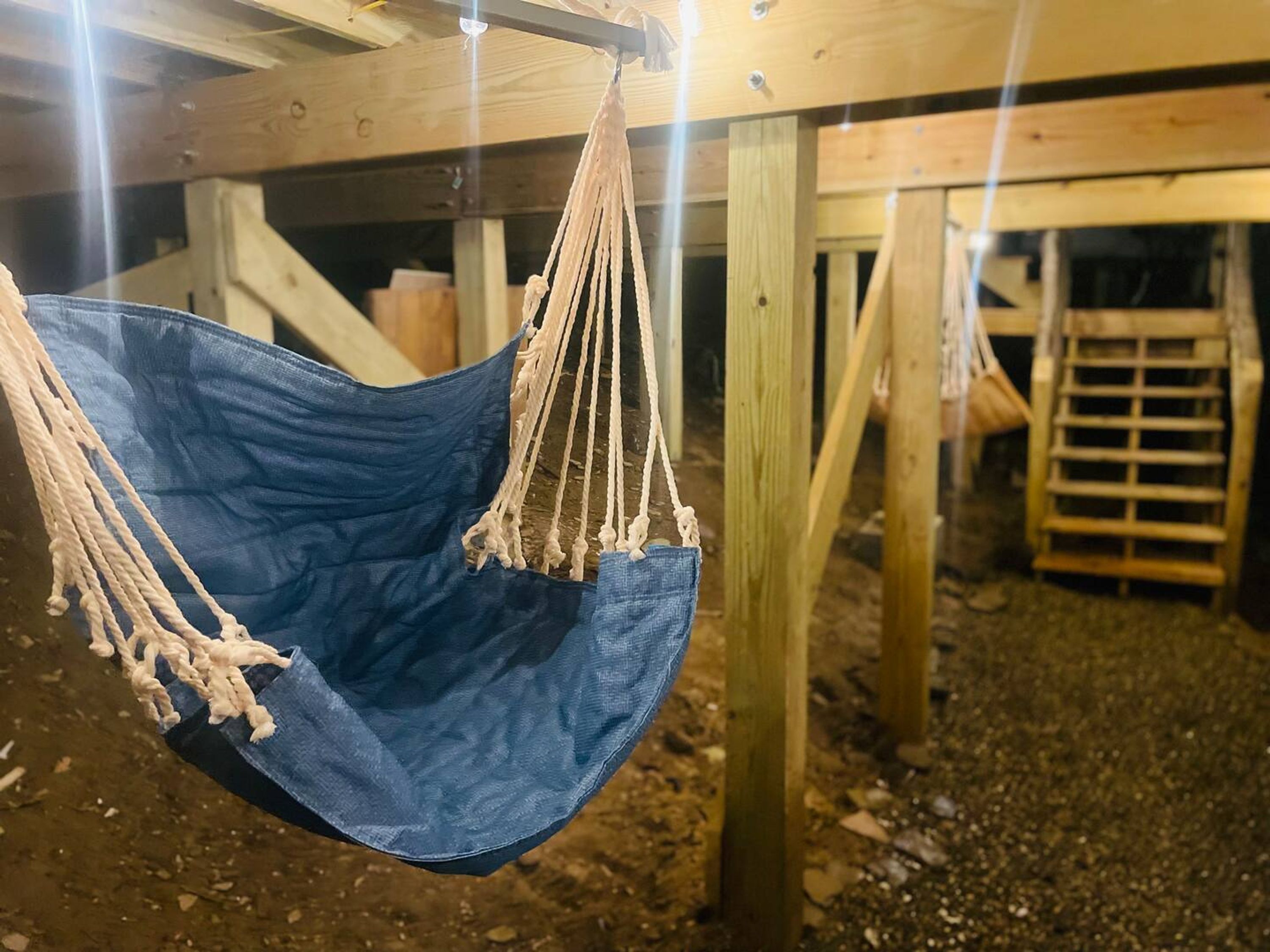 Uma rede pendurada em uma estrutura de madeira, possivelmente um alpendre ou terraço, com uma escada e vigas de madeira ao fundo.