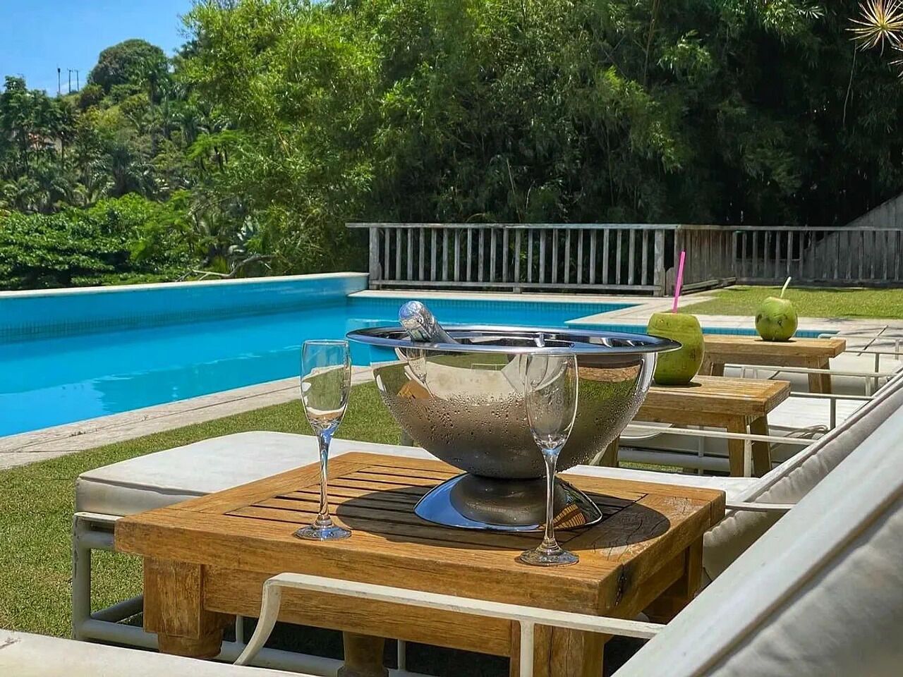 Uma área na piscina com espreguiçadeiras, uma mesa com uma tigela grande e copos de vinho, e uma cerca de madeira ao fundo.