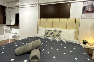 Quarto de hotel com cama, mesa de cabeceira com abajur, cadeira e vista para um edifício pela janela.