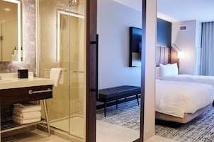 Quarto de hotel com banheiro, chuveiro, cama, um banco e uma TV.