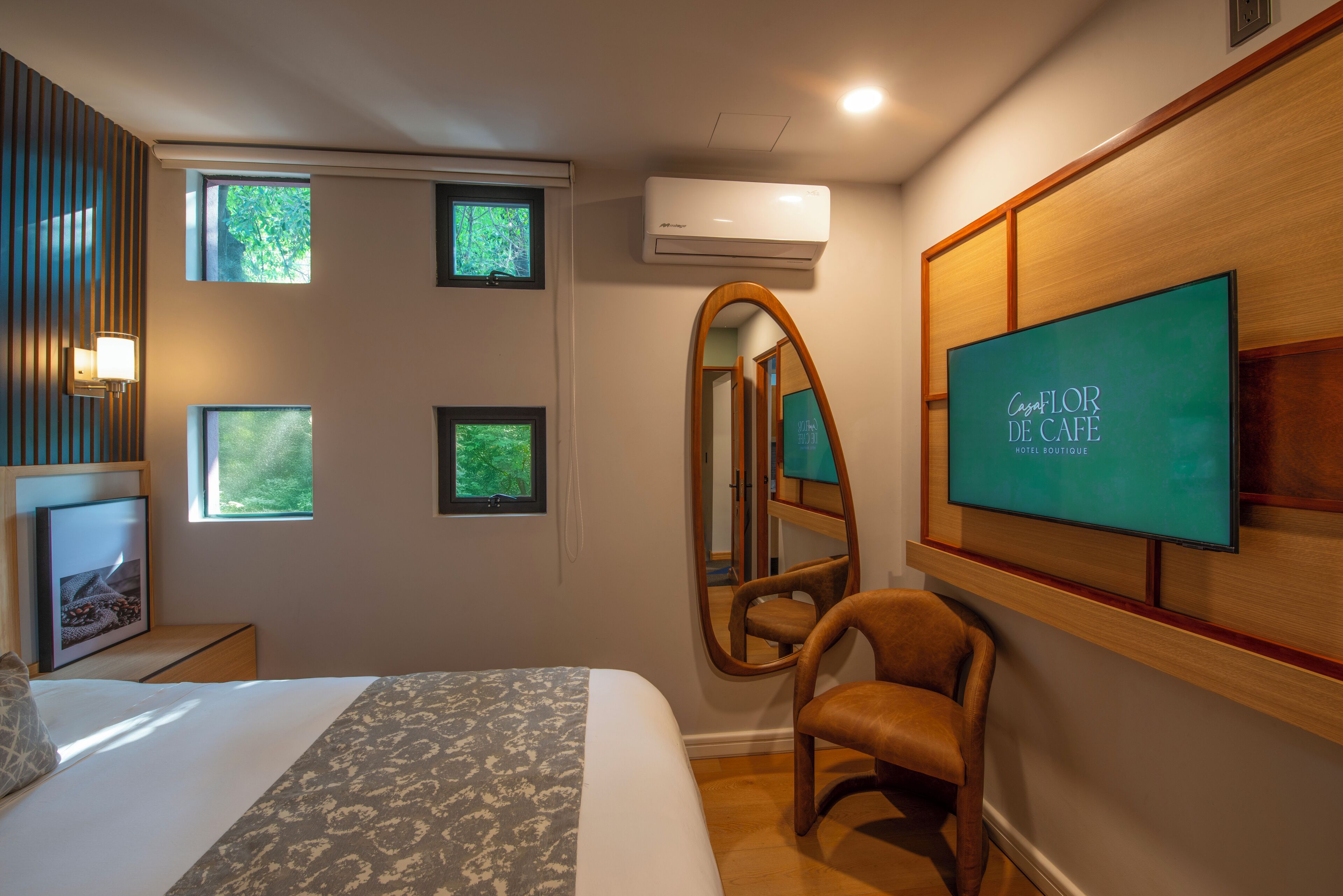 Quarto de hotel moderno com uma cama grande, uma cadeira de madeira, televisão e várias janelas.