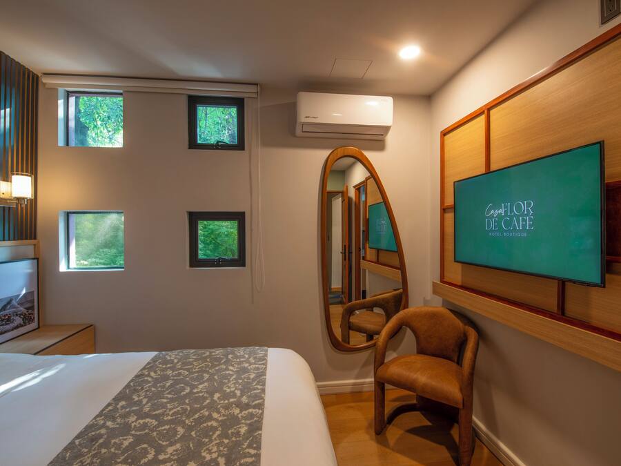 Quarto de hotel moderno com uma cama grande, uma cadeira de madeira, televisão e várias janelas.