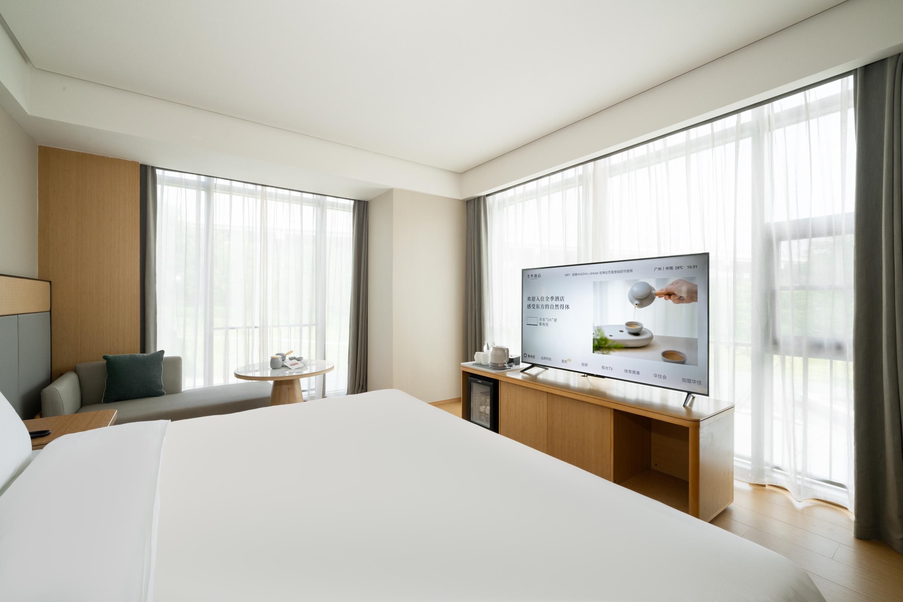 Quarto de hotel com uma cama grande, uma televisão sobre um suporte de madeira, uma mesinha com xícara e pires, e um sofá.