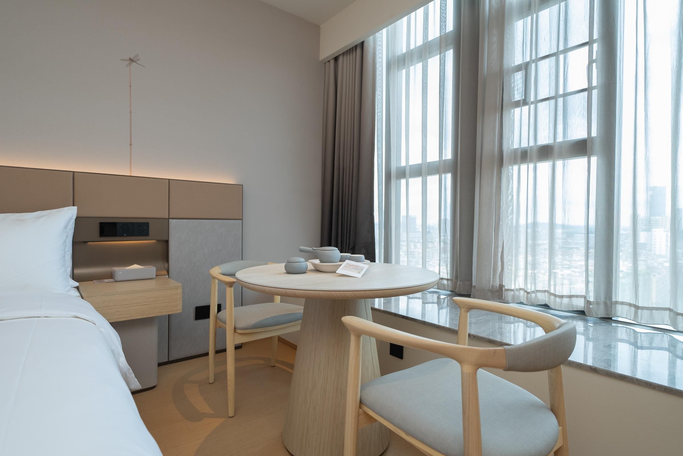 Quarto de hotel moderno com cama, mesa de cabeceira com abajur, mesa redonda com cadeiras e uma grande janela com cortinas.