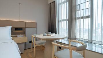 Quarto de hotel moderno com cama, mesa de cabeceira com abajur, mesa redonda com cadeiras e uma grande janela com cortinas.