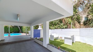 Uma área de varanda coberta com piscina, mobiliário de exterior branco e vista para um edifício branco ao fundo.