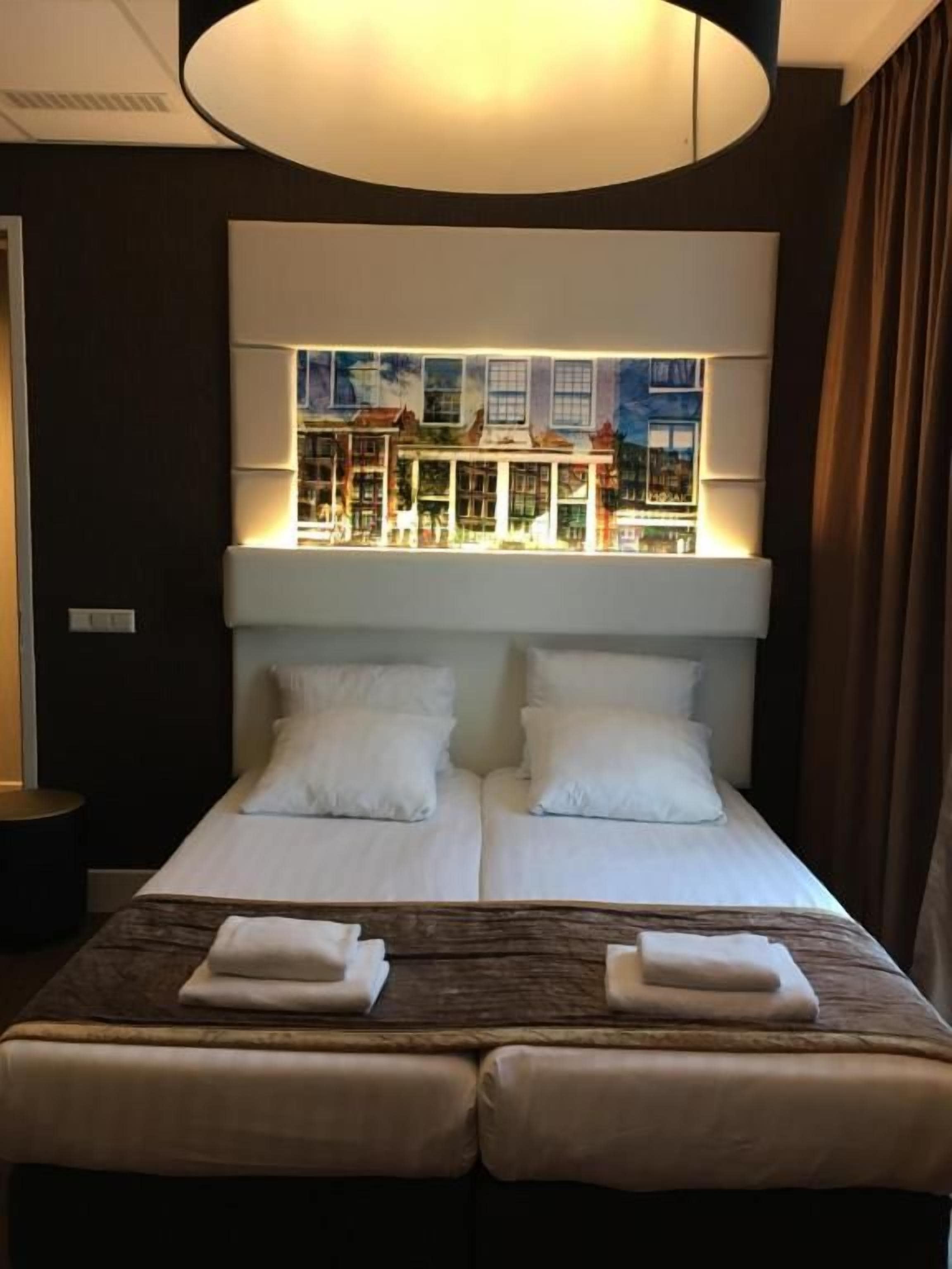 Uma cama de casal com cabeceira, duas almofadas, uma mesinha de cabeceira e uma janela com vista para edifícios.