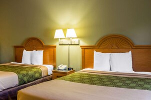 Quarto de hotel com duas camas, cada uma com colcha estampada, uma mesinha de cabeceira com abajur e luminária na parede.