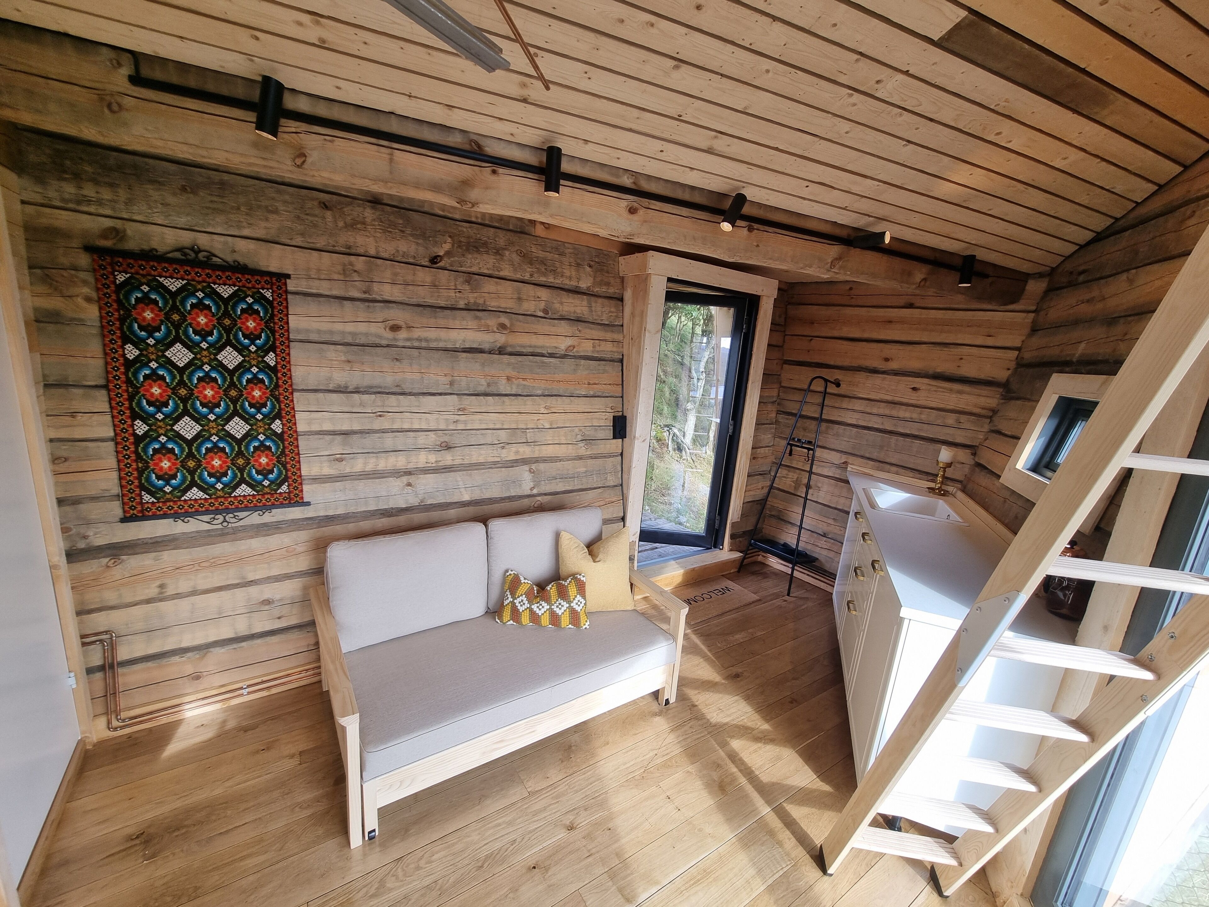 Interior de uma cabana com um sofá, uma janela, uma escada de madeira e um tapete fixado na parede.