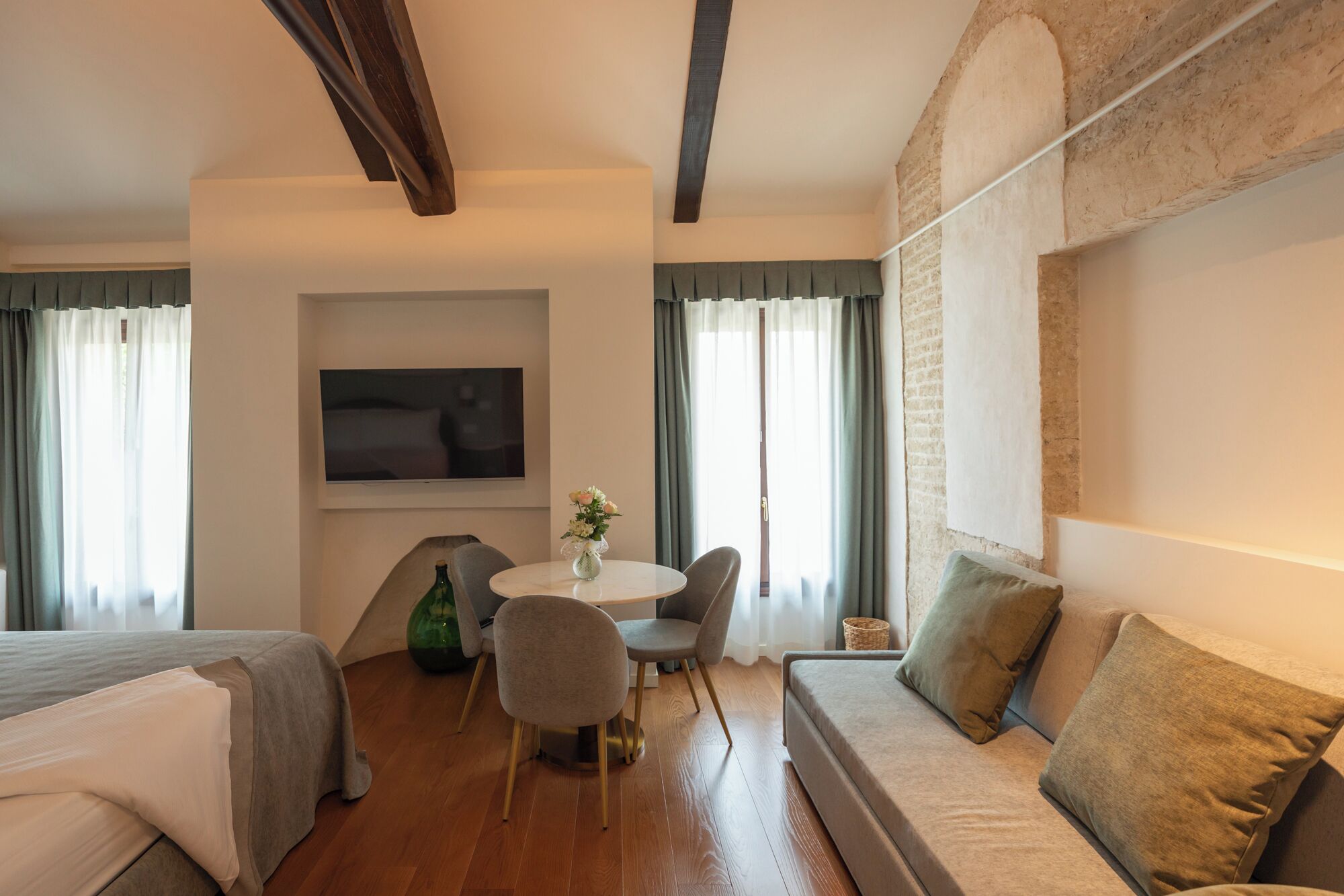 Quarto de hotel com cama, sofá, mesa de jantar com cadeiras, televisão na parede e janela com cortinas.