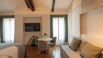 Quarto de hotel com cama, sofá, mesa de jantar com cadeiras, televisão na parede e janela com cortinas.