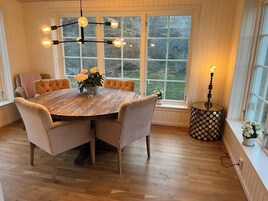 Uma sala de jantar com uma mesa redonda de madeira, seis cadeiras estofadas, um lustre e uma janela com vista para árvores.