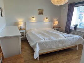 Quarto bem arrumado com uma cama grande, duas mesas de cabeceira com abajures, um criado-mudo de madeira, uma cômoda branca e uma janela com cortinas.