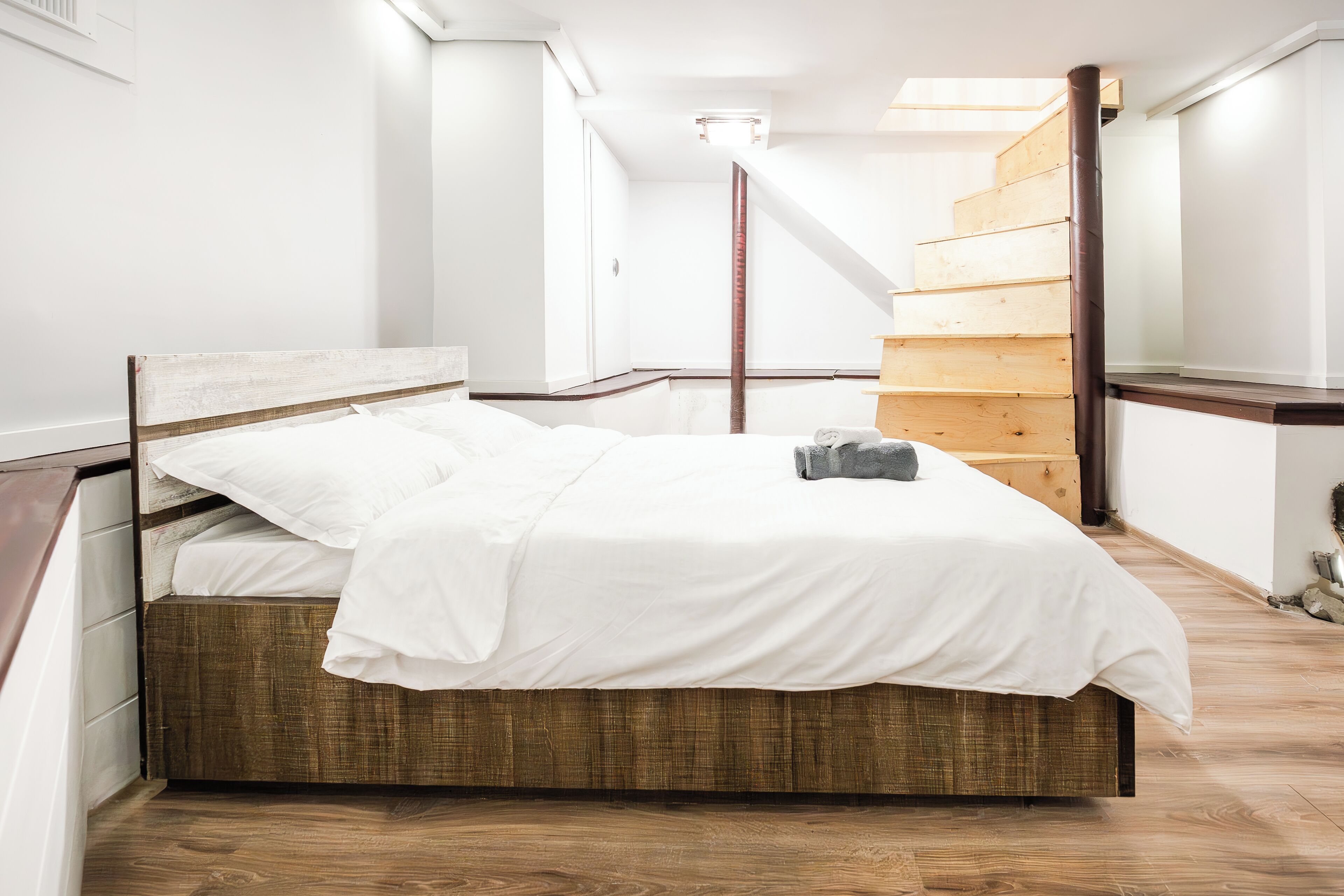 Quarto com estrutura de cama de madeira, roupa de cama branca e escada de madeira ao fundo.