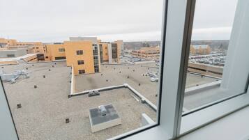 Vista da janela sobre um parque de estacionamento e edifícios, incluindo uma construção de cobertura plana e um edifício central com fachada de vidro.