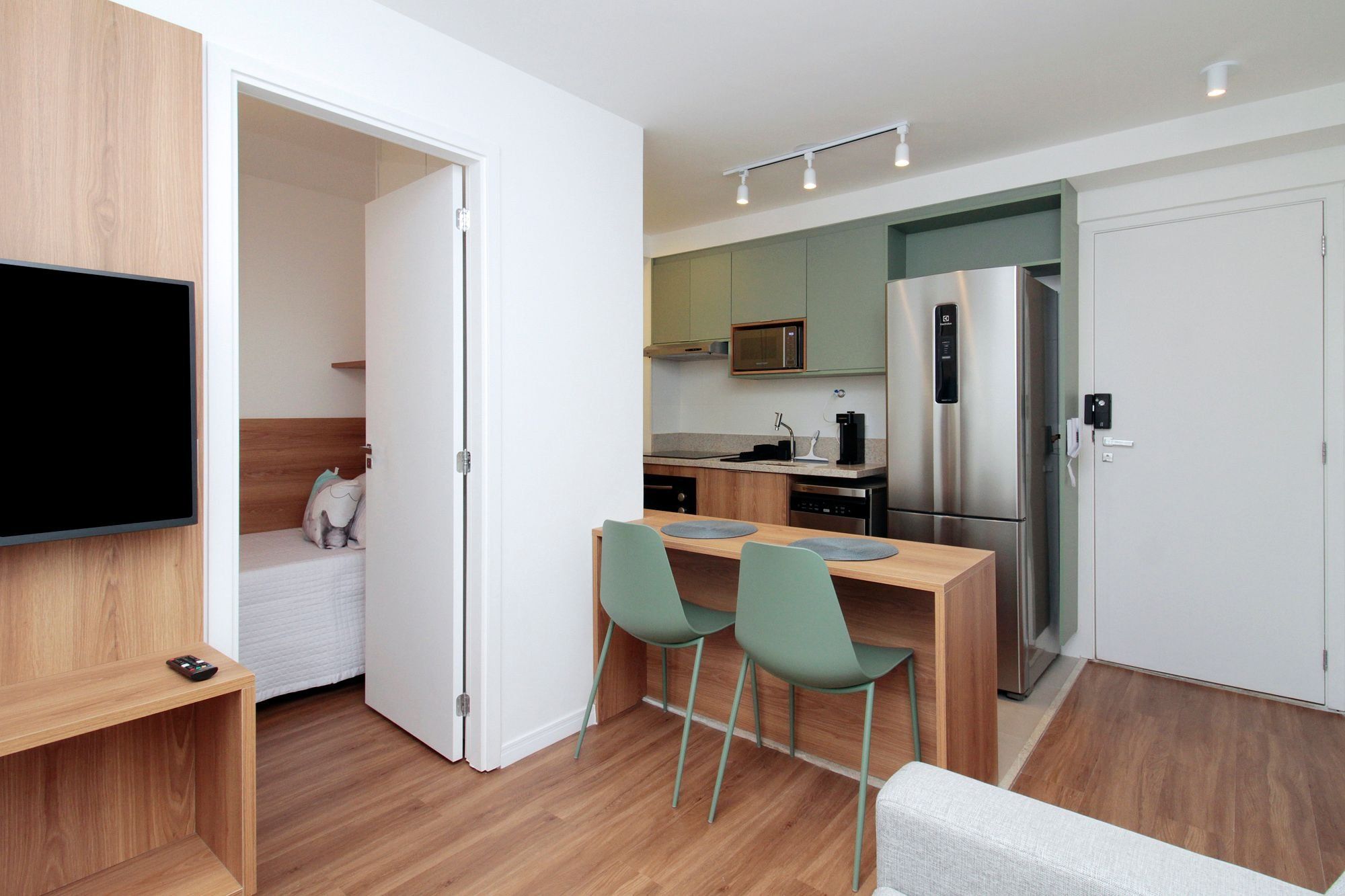 Um espaço compacto com cozinha compacta, área de jantar com duas cadeiras, TV fixada em um painel de madeira e uma cama com almofadas.