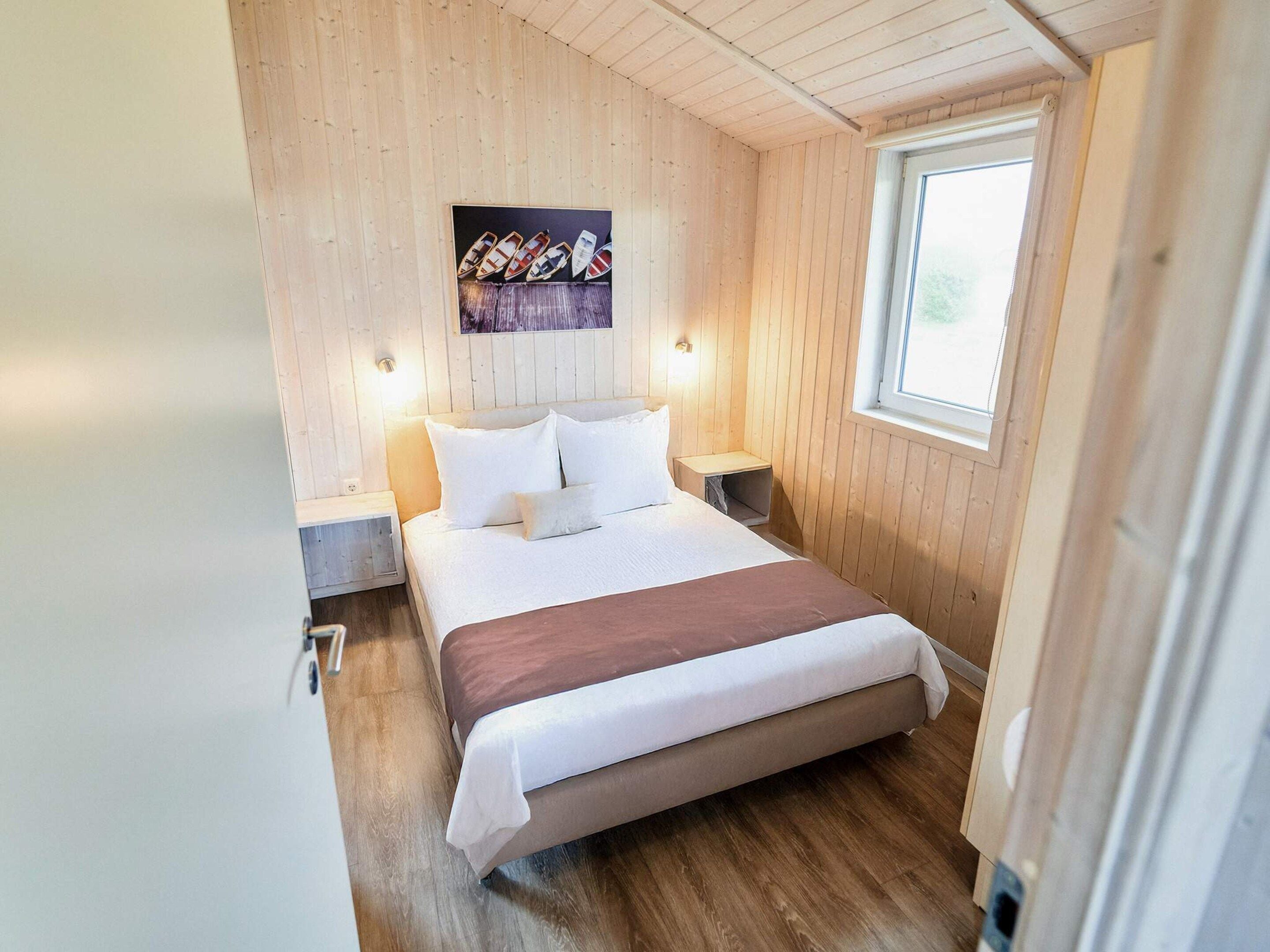 Um quarto pequeno e acolhedor com teto de madeira, uma cama de solteiro com roupa de cama branca, uma mesa de cabeceira e uma janela.