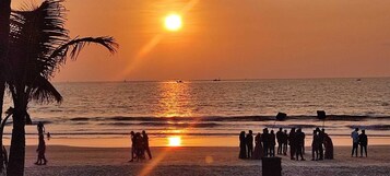 Cena de praia com o sol se pondo sobre o oceano, palmeiras e silhuetas de pessoas na areia.