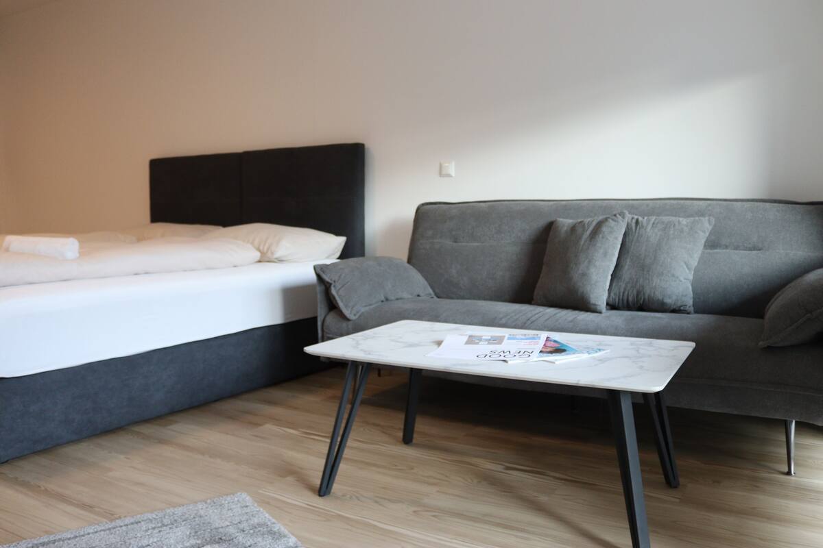Quarto moderno com uma cama, um sofá cinzento e uma pequena mesa com revistas.